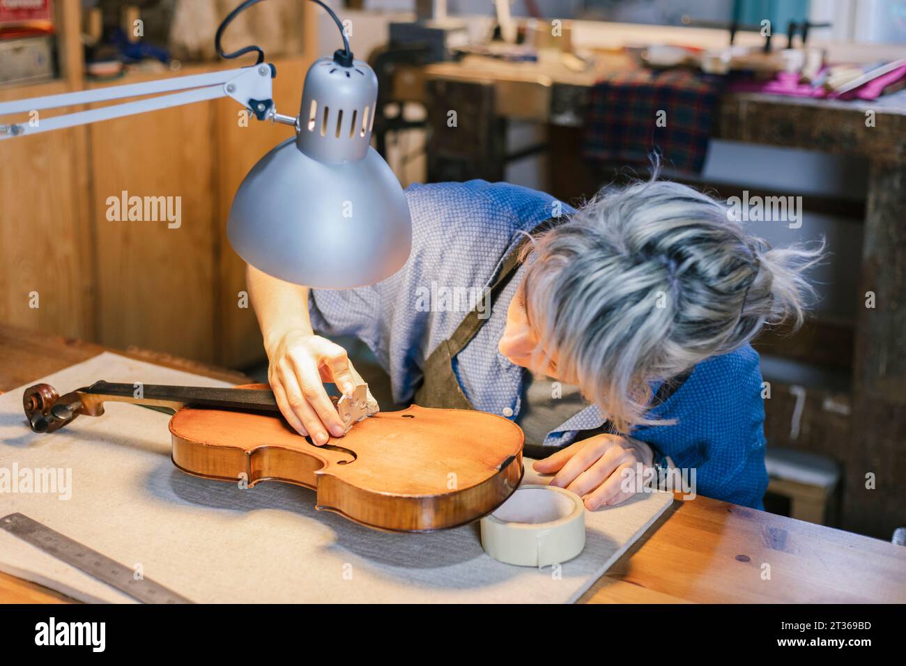 Instrument maker arranging bridge part on violin at desk in workshop ...