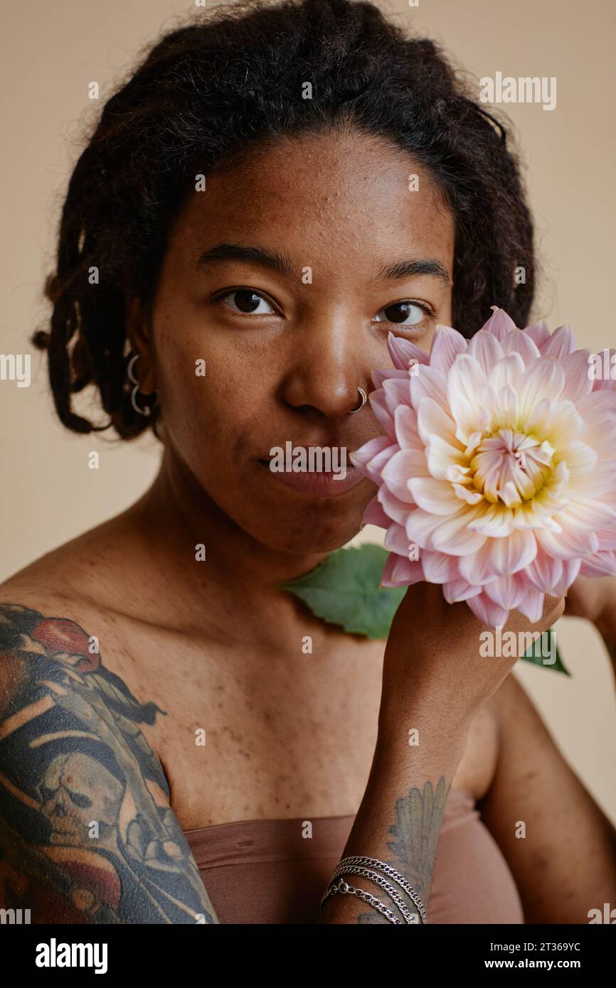 Young woman with tattoo and acne scars holding flower against brown ...