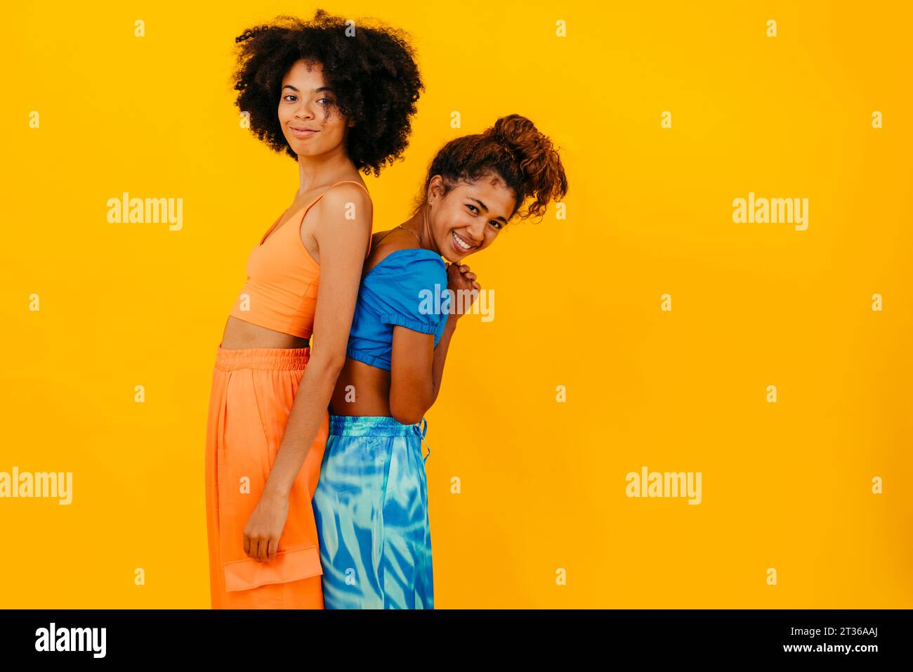 Smiling young friends standing back to back against yellow background ...