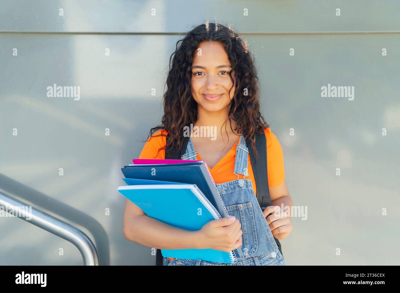 Smiling student holding spiral notebooks in front of wall at campus ...