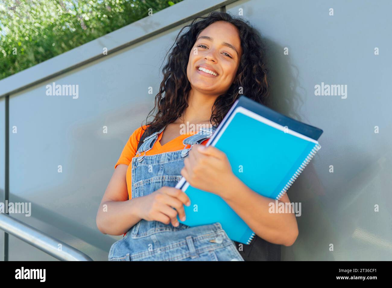 Happy student holding spiral notebooks and leaning on wall in campus ...