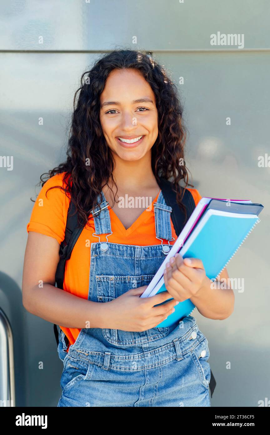 Happy student holding spiral notebooks in front of wall at campus Stock ...