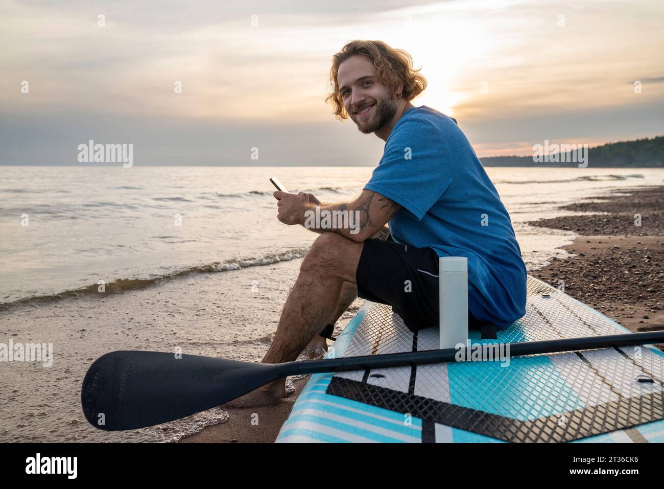 Smiling man holding smart phone sitting on paddleboard at beach Stock ...