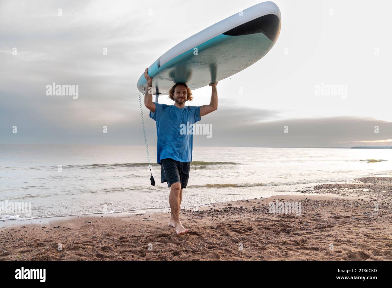 Smiling man carrying paddleboard and walking at beach Stock Photo - Alamy