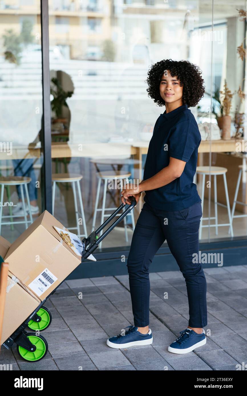 Smiling delivery woman pushing goods in cart near cafe window Stock ...