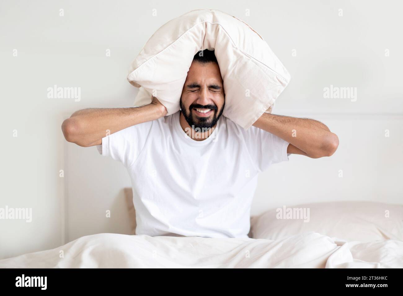 Stressed Young Indian Man Sitting In Bed And Covering Ears With Pillow ...