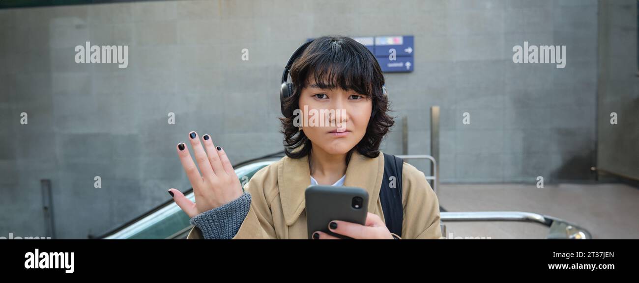 Portrait of asian girl in headphones, looks complicated at smartphone ...