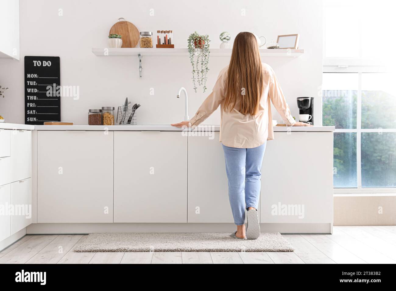 Back view of young woman standing near white counters in modern kitchen ...