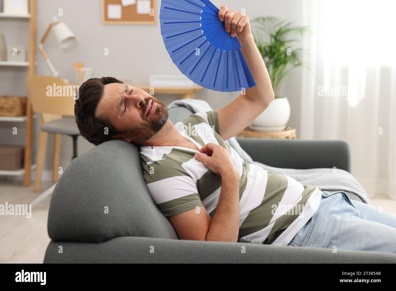 Bearded man waving blue hand fan to cool himself on sofa at home Stock ...