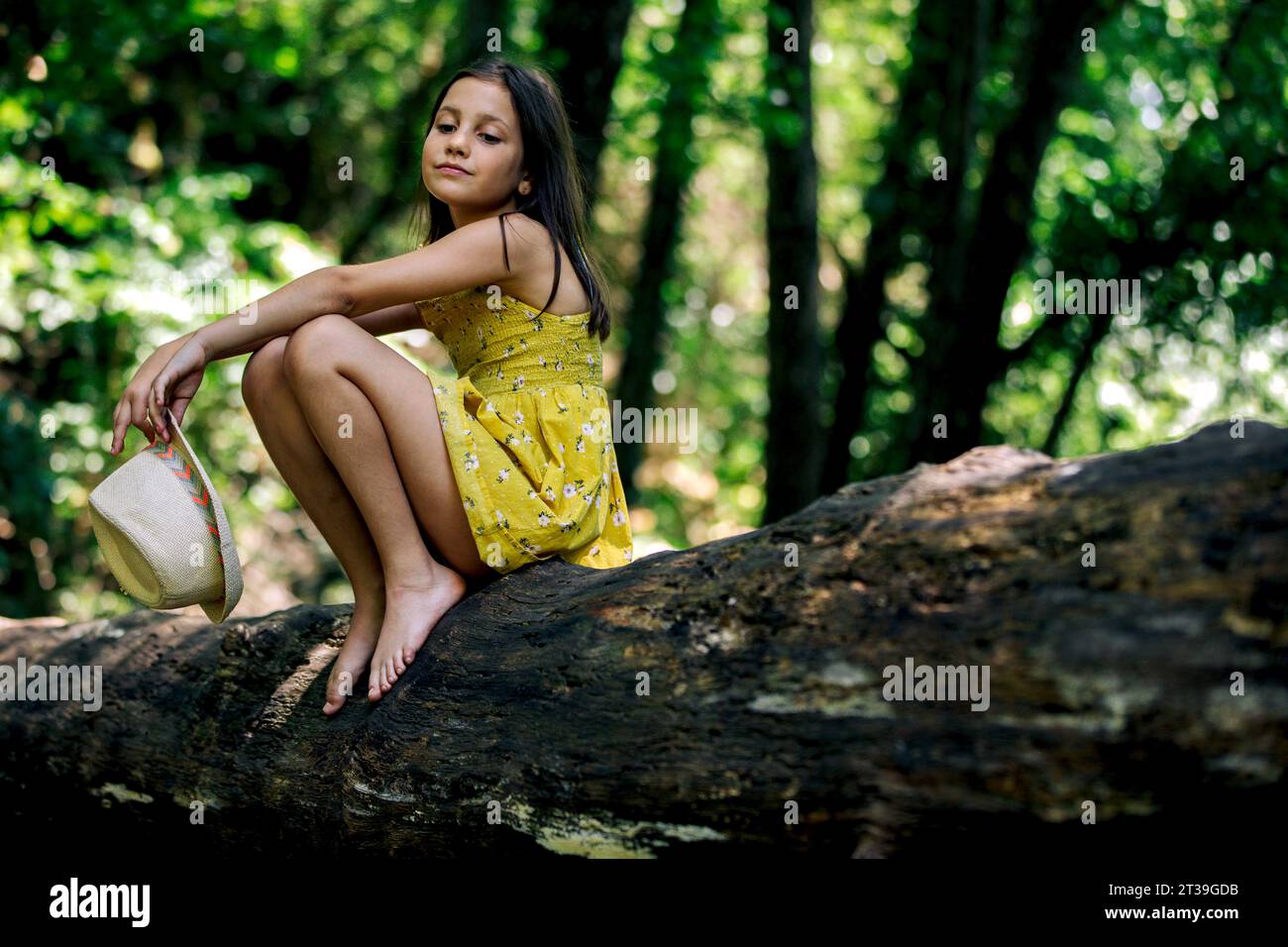 Side view of pensive preteen girl in dress holding hat while sitting on ...
