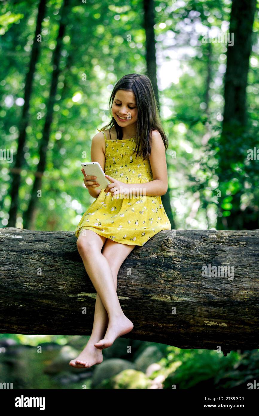 Smiling preteen girl wearing dress sitting on tree trunk while taking selfie with smartphone in ...