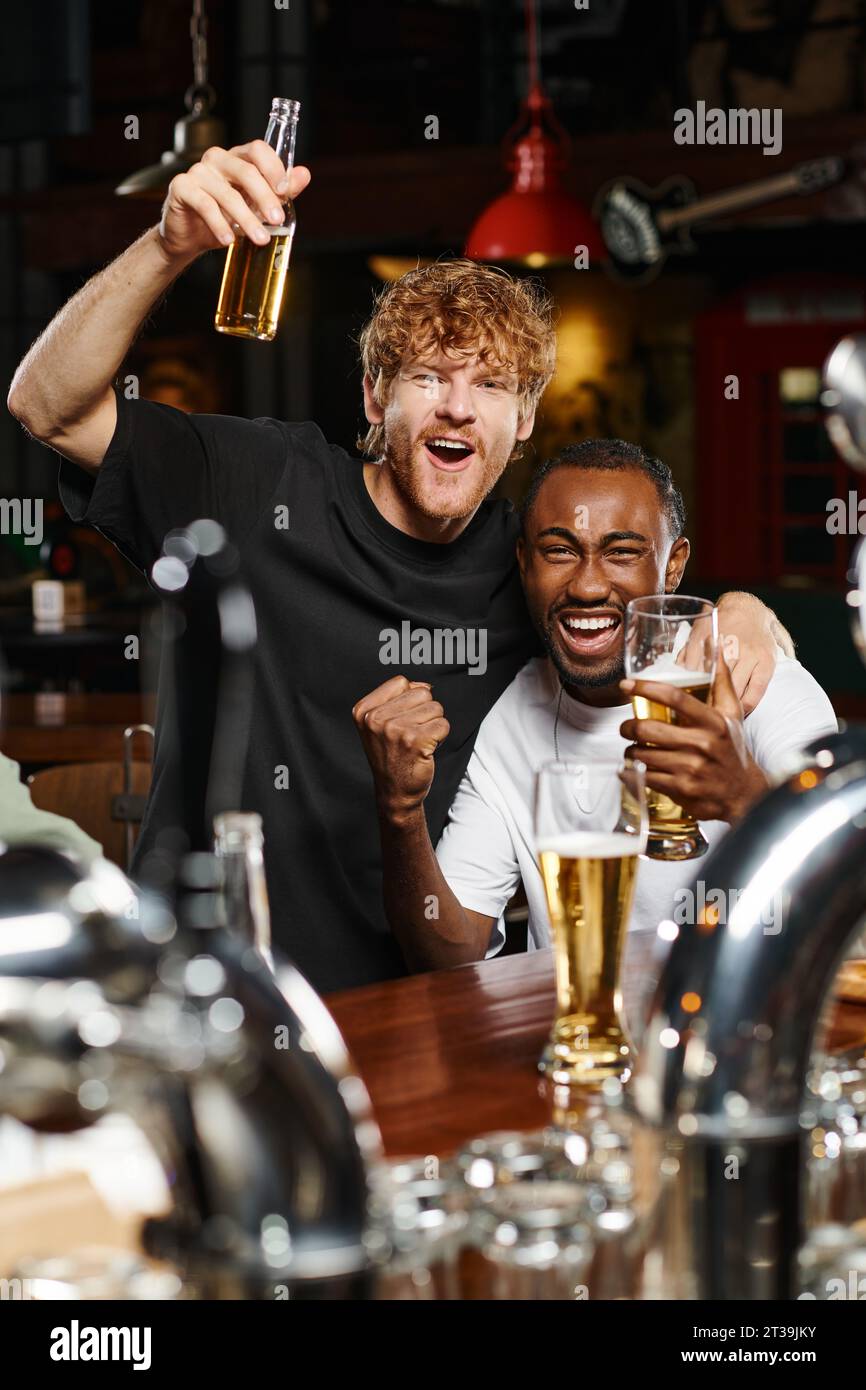 excited interracial friends hugging and raising glasses of beer while ...