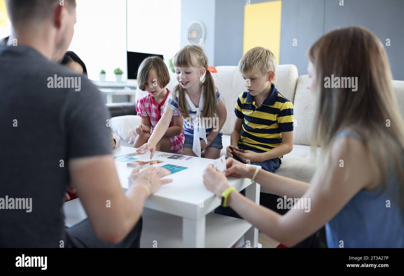 Adults and children are sitting around table on which playing cards are ...