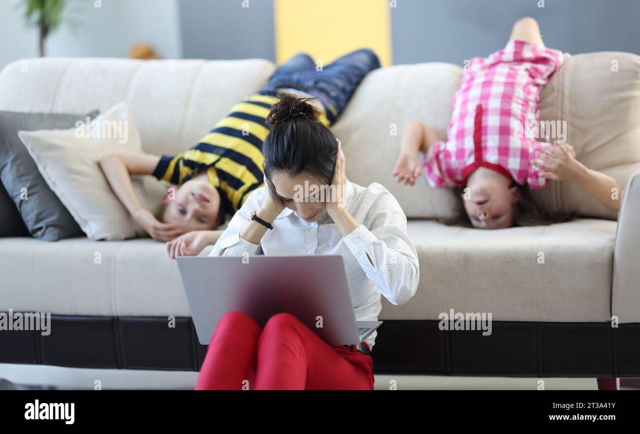 Woman sits on floor with a laptop and covers her ears with her hands. behind her on couch are ...