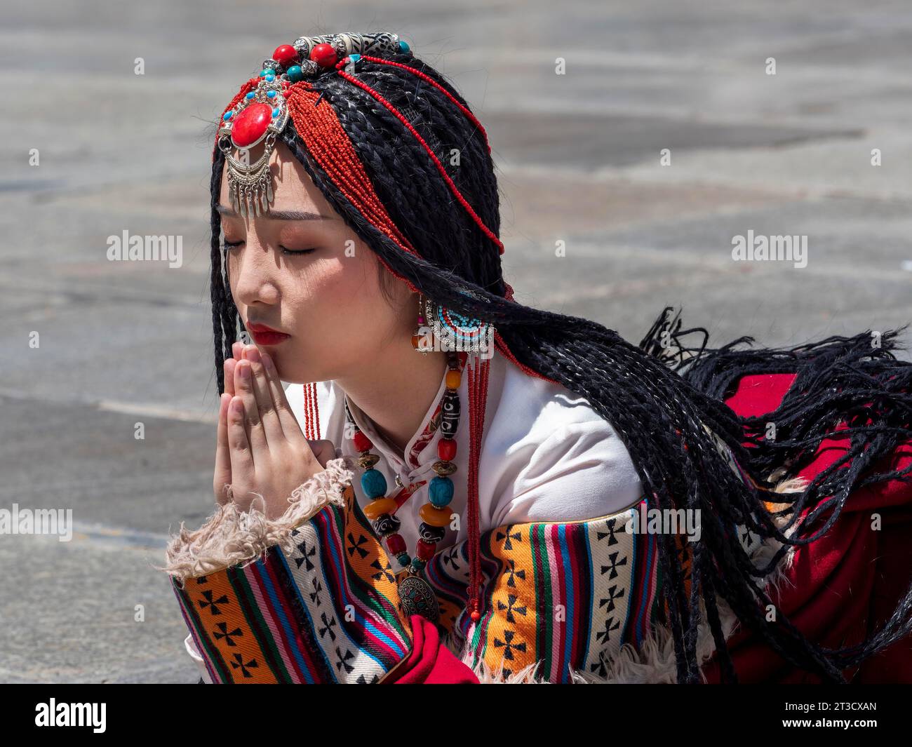 Tibetan young woman with festive hair ornaments and necklaces praying on the floor, exotic ...