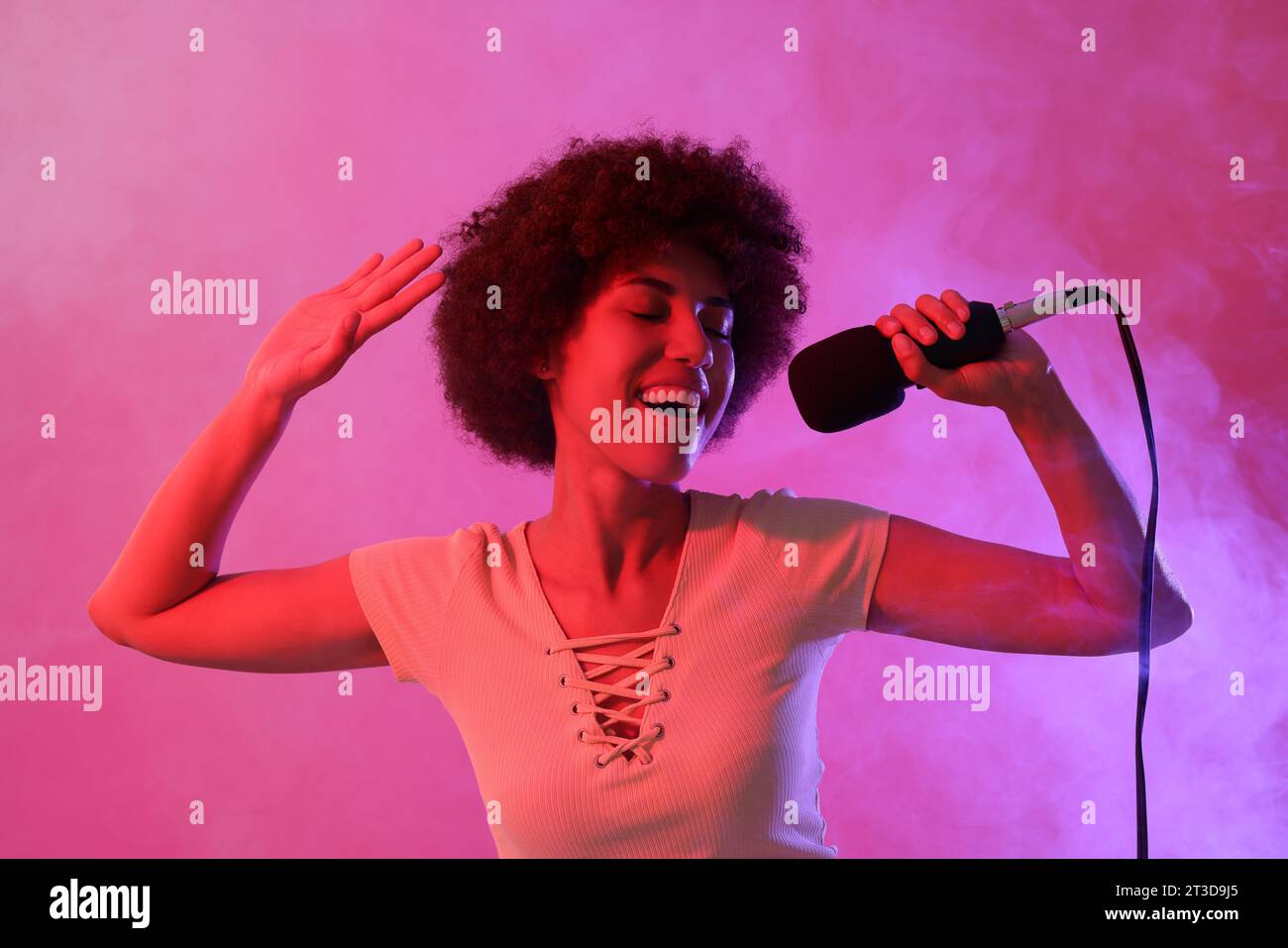 Curly young woman with microphone singing on pink background. Color ...