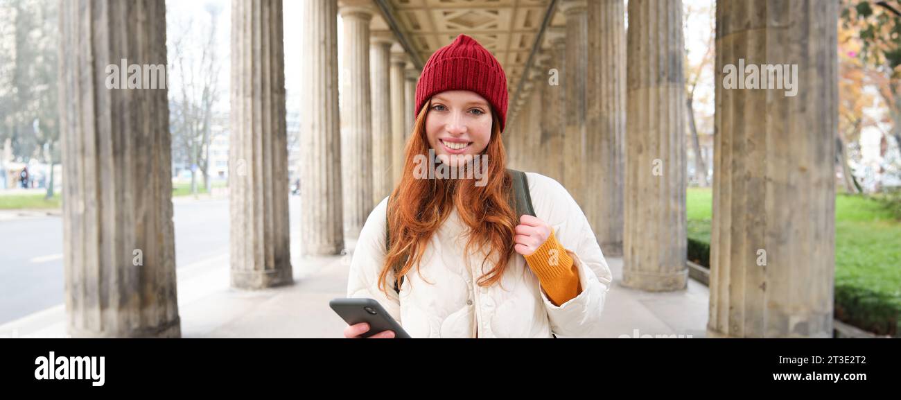 Happy young female model stands on street and holds mobile phone, uses ...