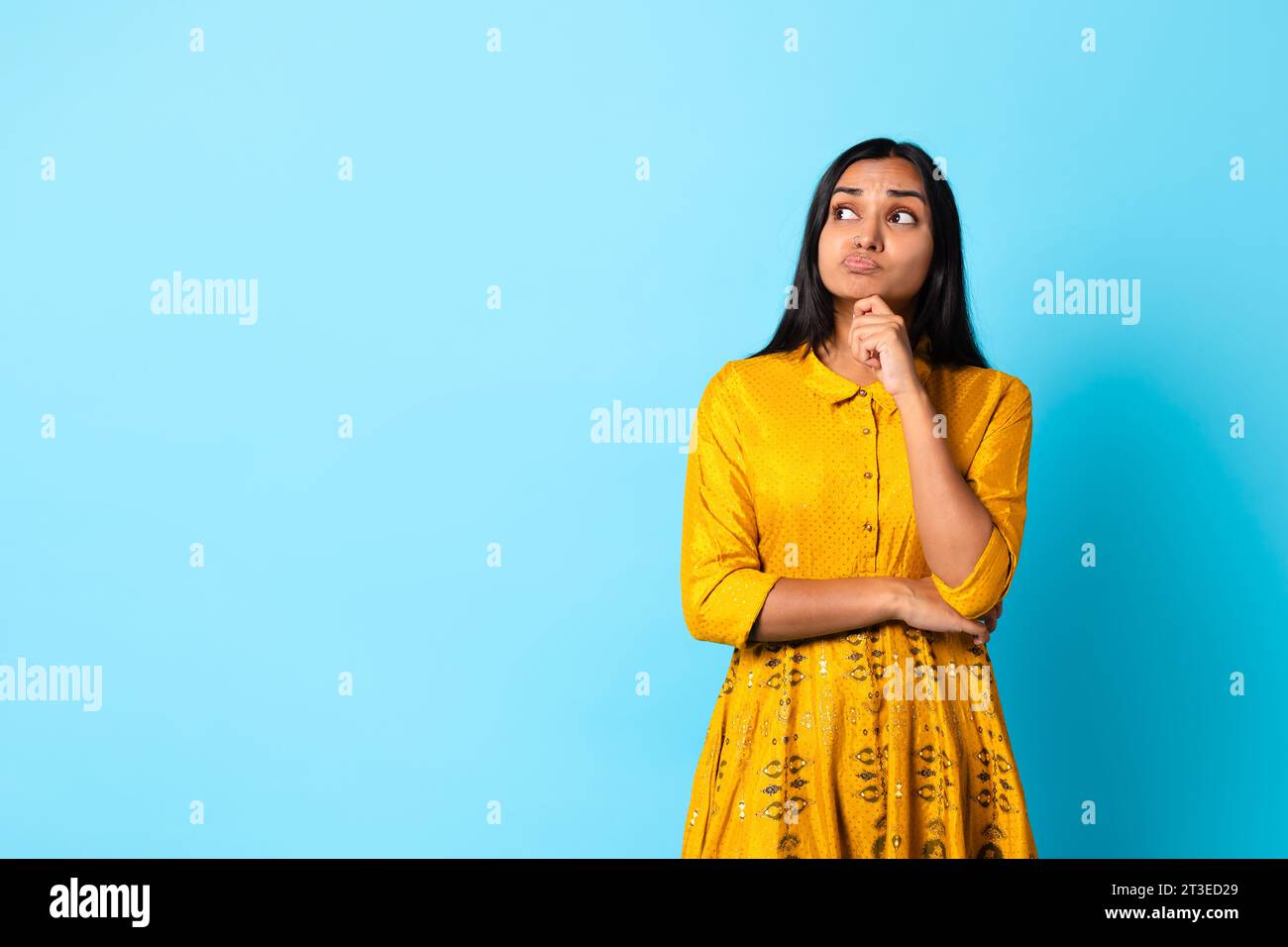 Thoughtful young Indian lady in traditional yellow dress, blue backdrop ...