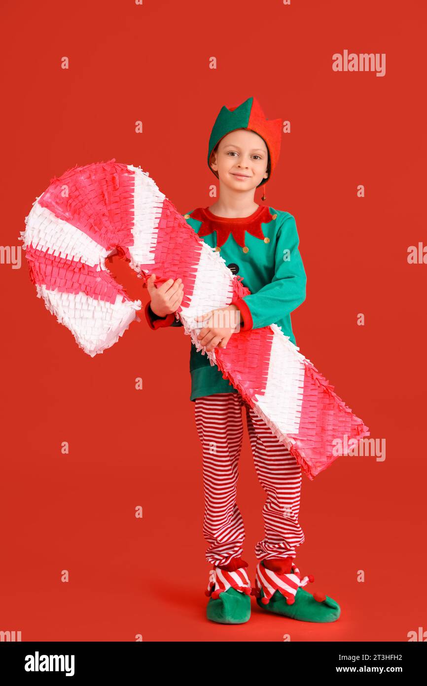 Cute little boy in elf costume with candy cane pinata on red background ...
