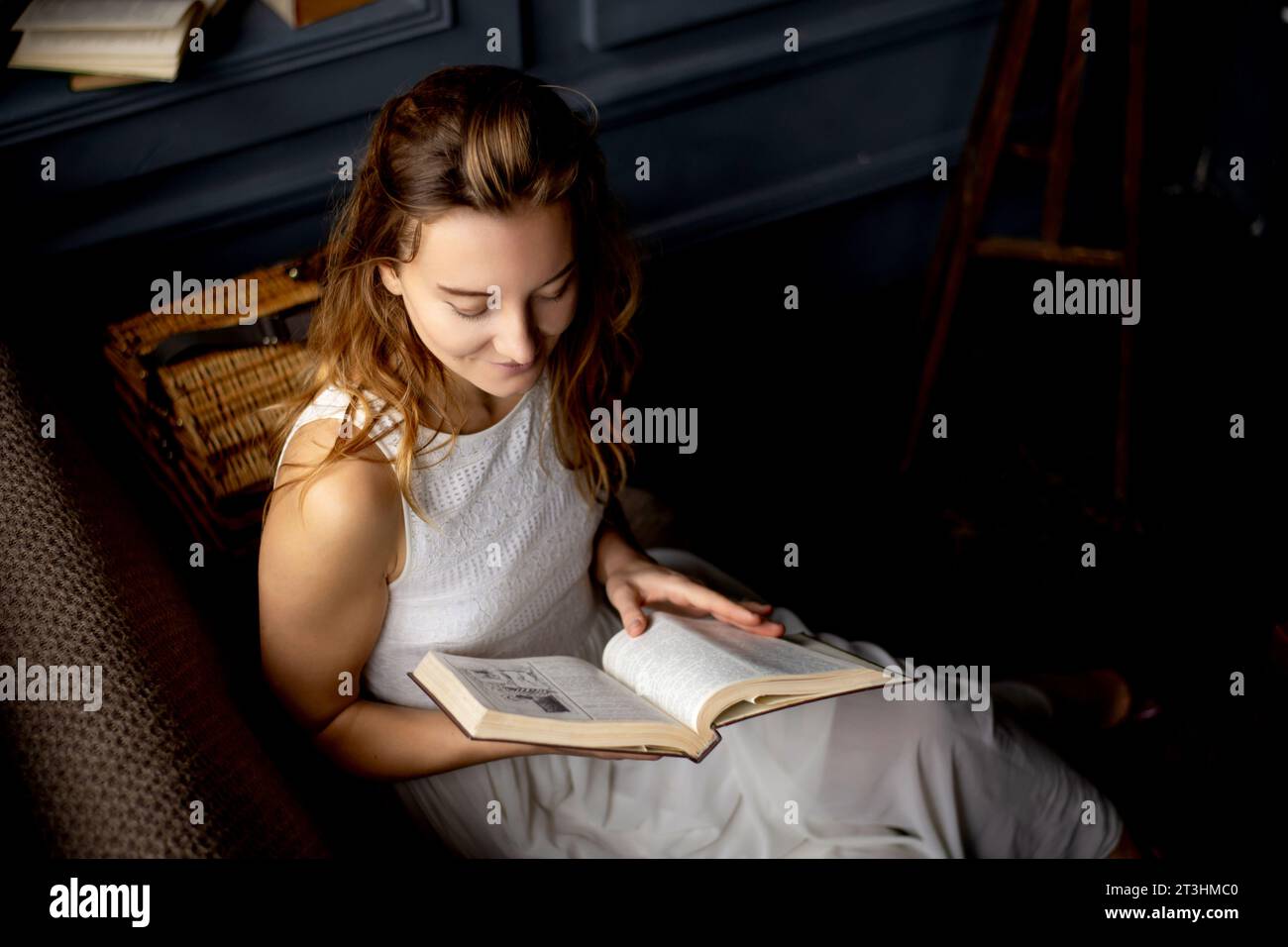 In the soft glow, a young woman with flowing hair smiles as she reads a book, seated on the sofa ...