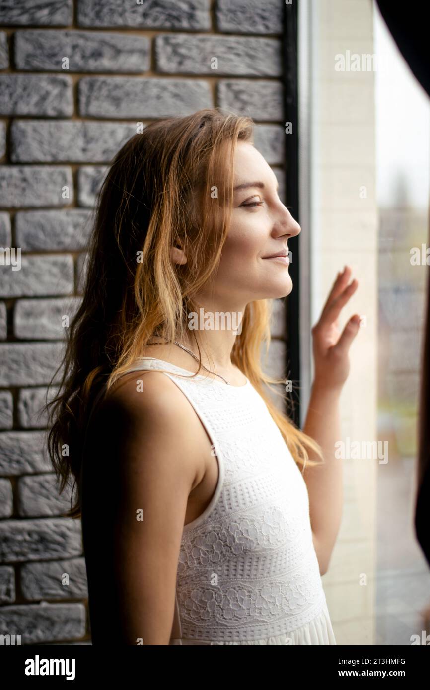 A young lady, dressed in ethereal white, beams near the vast window ...