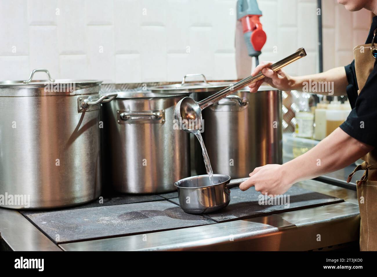 Hands of young male chef in apron pouring hot water into ladle while ...