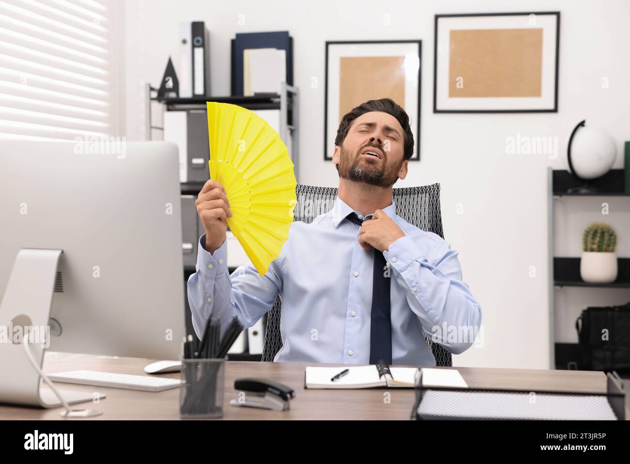 Bearded businessman waving yellow hand fan to cool himself at table in ...