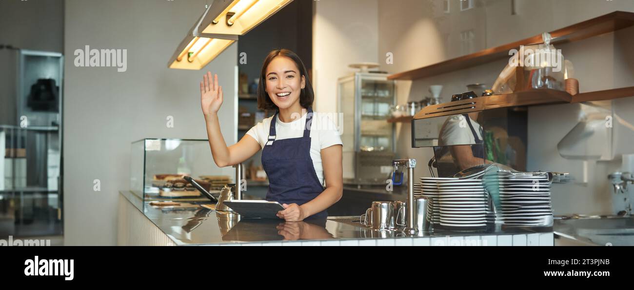 Cheerful asian girl barista, bartender behind counter raising arm and ...