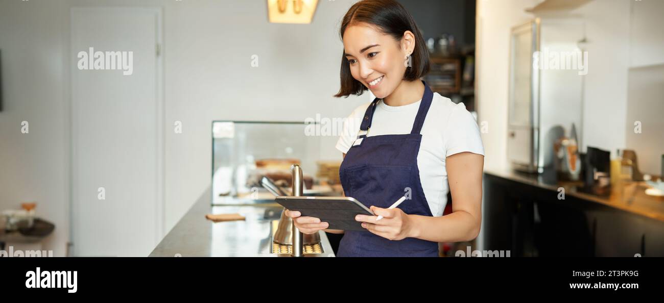 Small cafe business. Smiling asian girl barista in apron, using tablet ...