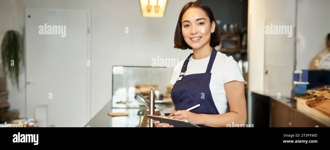 Small cafe business. Smiling asian girl barista in apron, using tablet ...