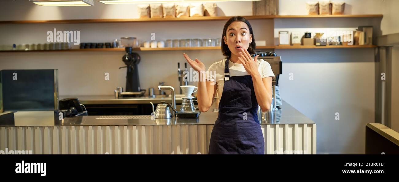 Image of excited asian female barista, looking surprised, pointing at ...