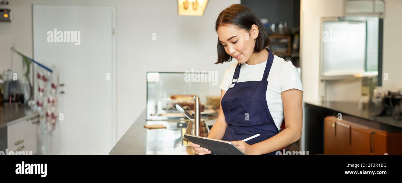 Small cafe business. Smiling asian girl barista in apron, using tablet ...