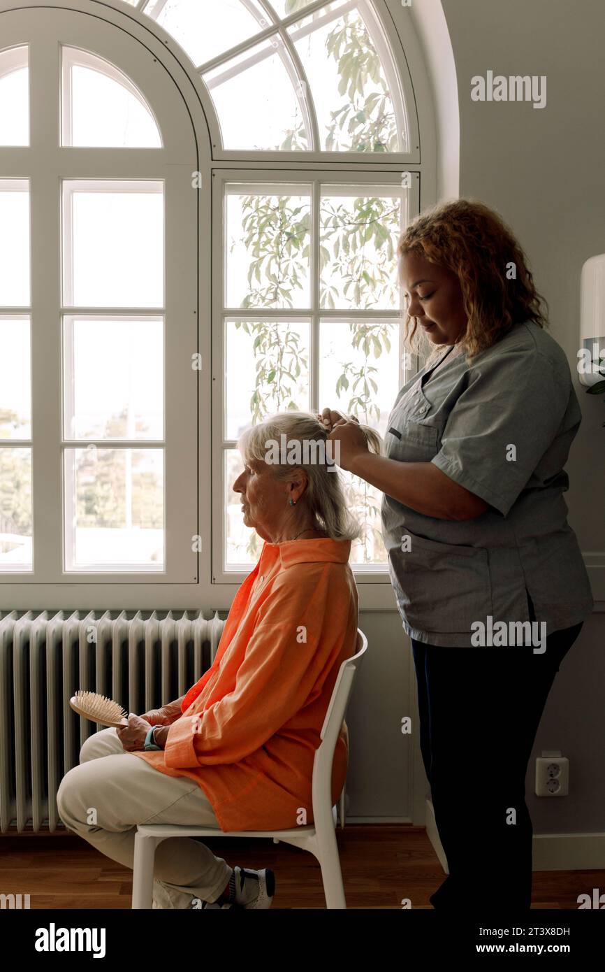 Side view of young female caregiver braiding senior woman's hair at ...