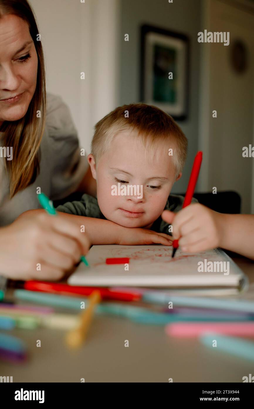 Boy with down syndrome drawing in book by mother at dining table Stock ...