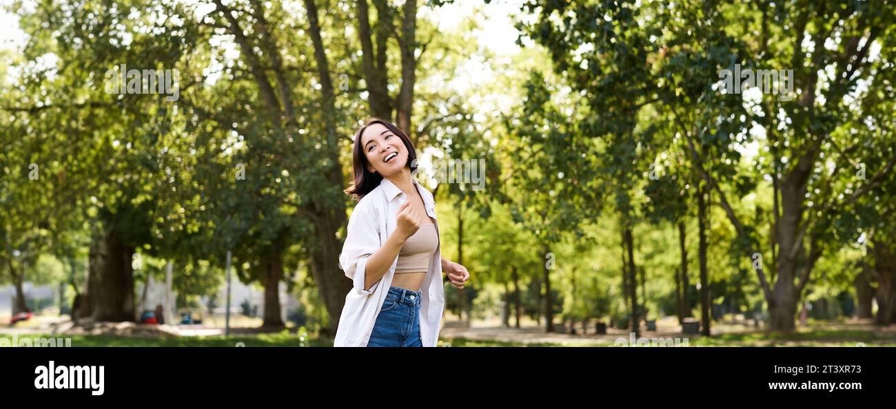 Happy young asian woman walking alone, dancing and singing in park ...