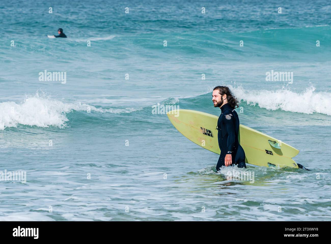A male surfer carrying his surfboard wading out to sea at Fistral in ...