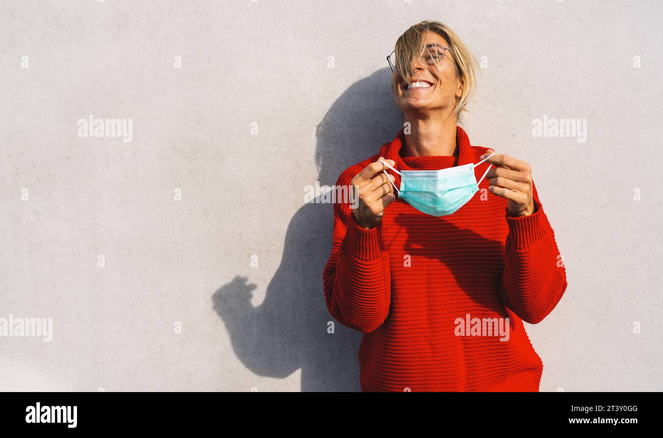 A joyful woman in a bright red sweater removing a light blue face mask ...