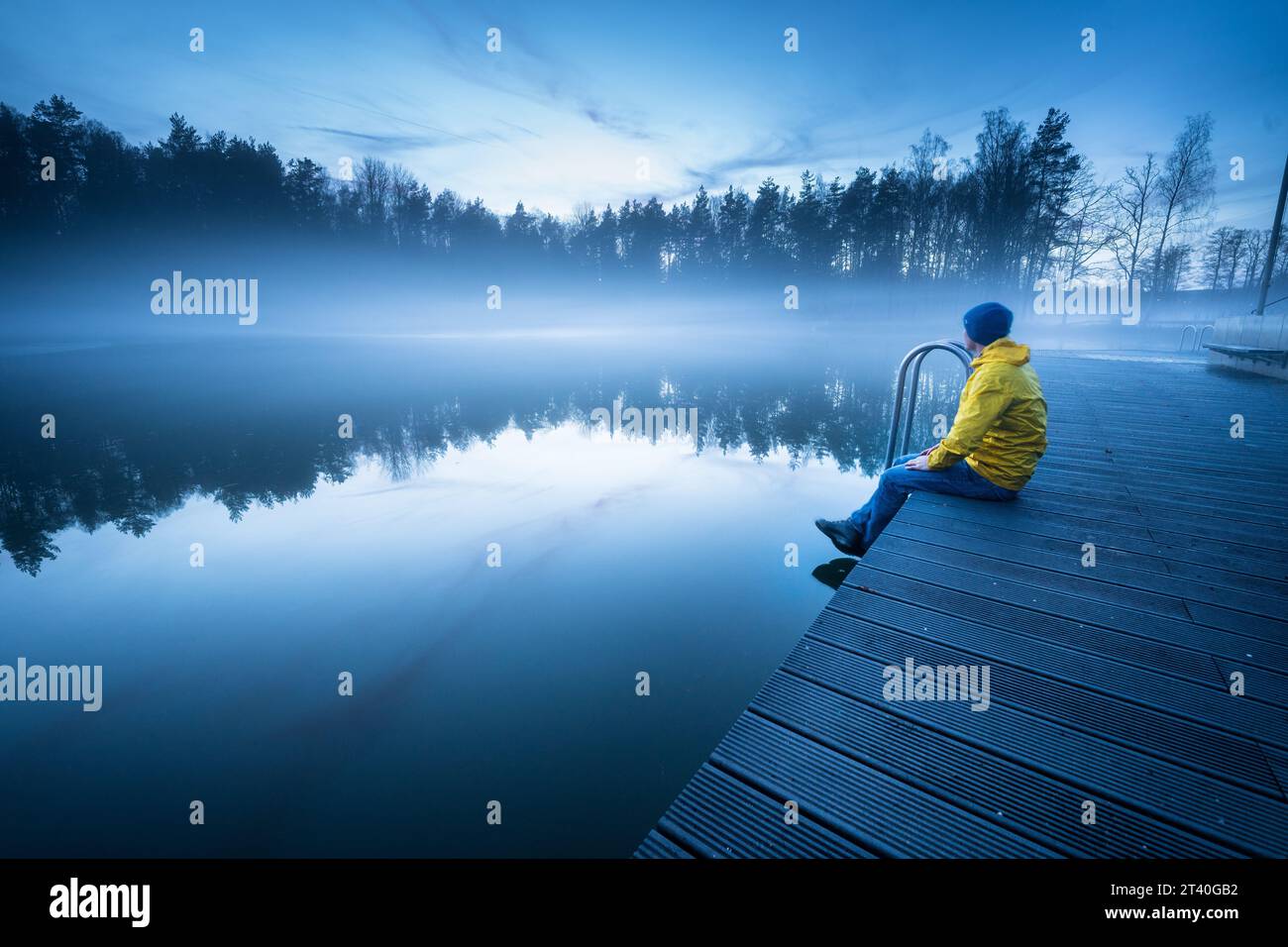 A young man in outdoor attire sits on a pier overlooking a tranquil ...
