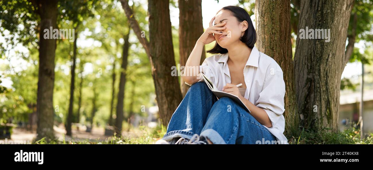 Sad girl crying while writing in her diary, sitting alone under tree in ...