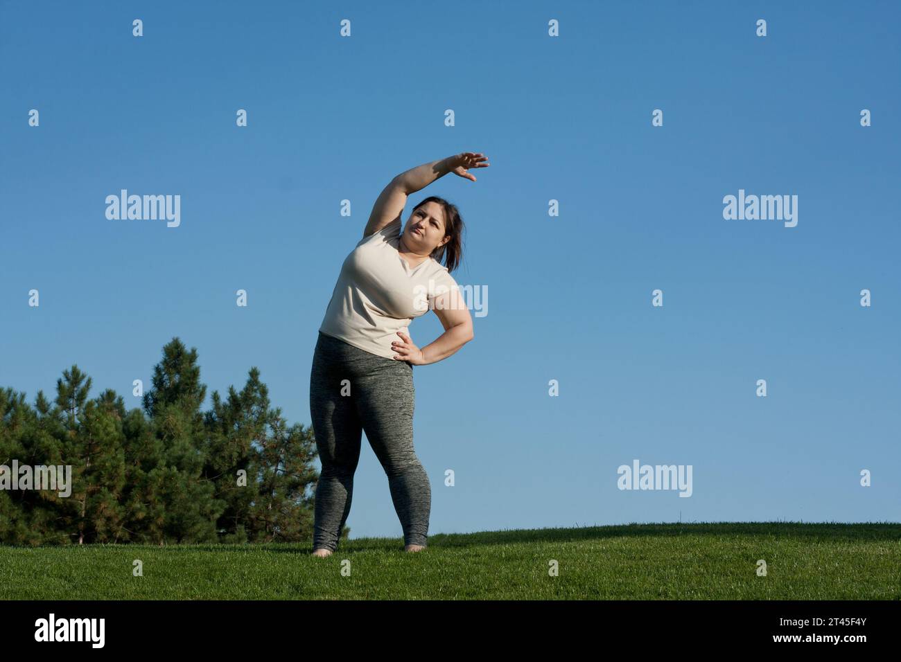 overweight middle-aged woman does exercises in park standing on grass ...