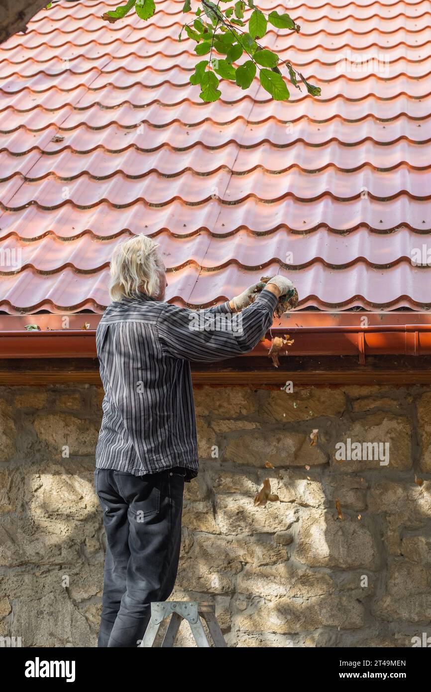A mature man removes leaves and debris from the gutter of his house ...