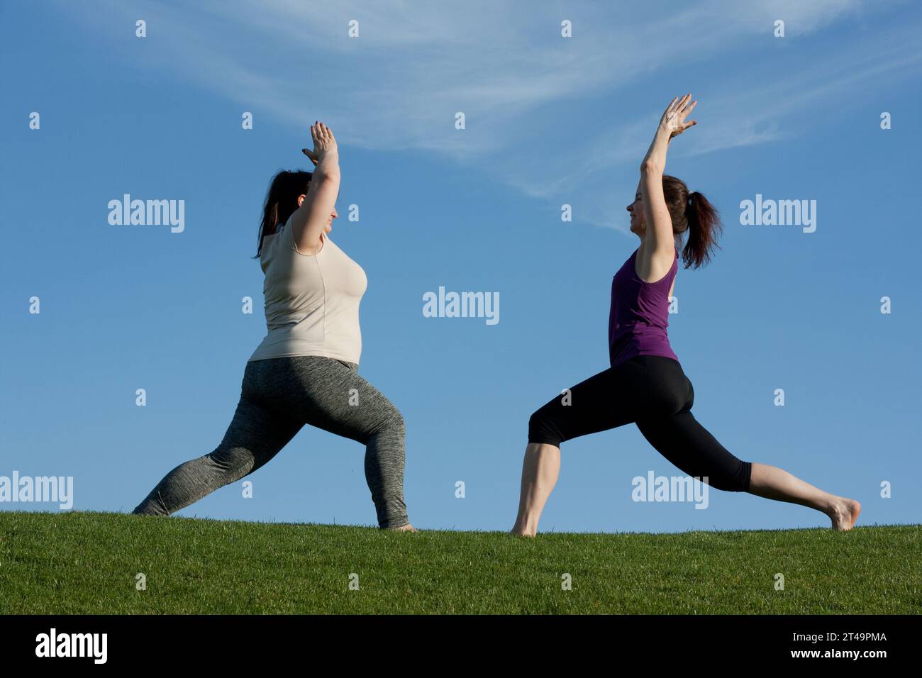 Two middle-aged women practice yoga in city park in warrior pose, blue sky background. Healthy ...