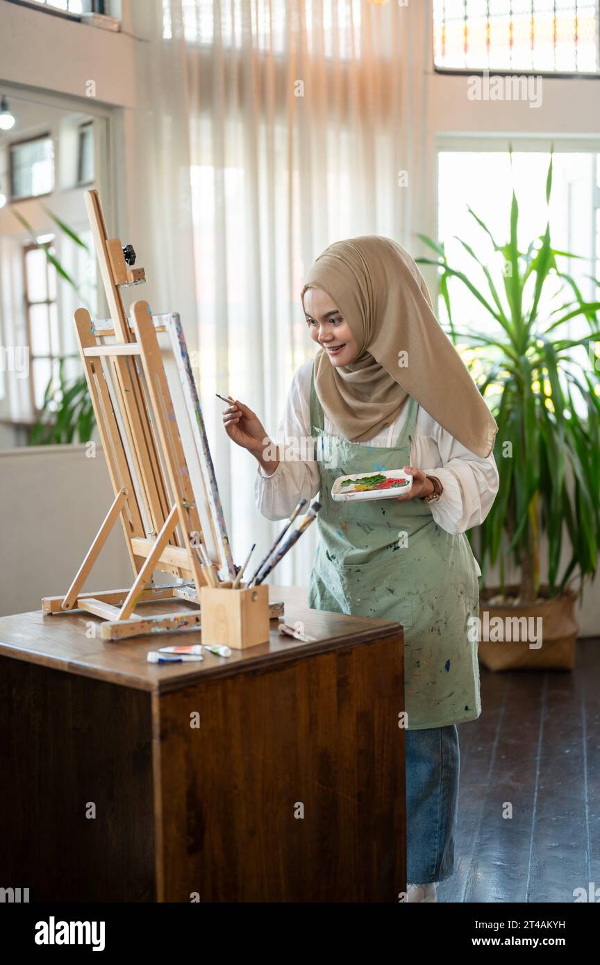 A happy and creative Asian-Muslim female artist in a hijab and apron is ...