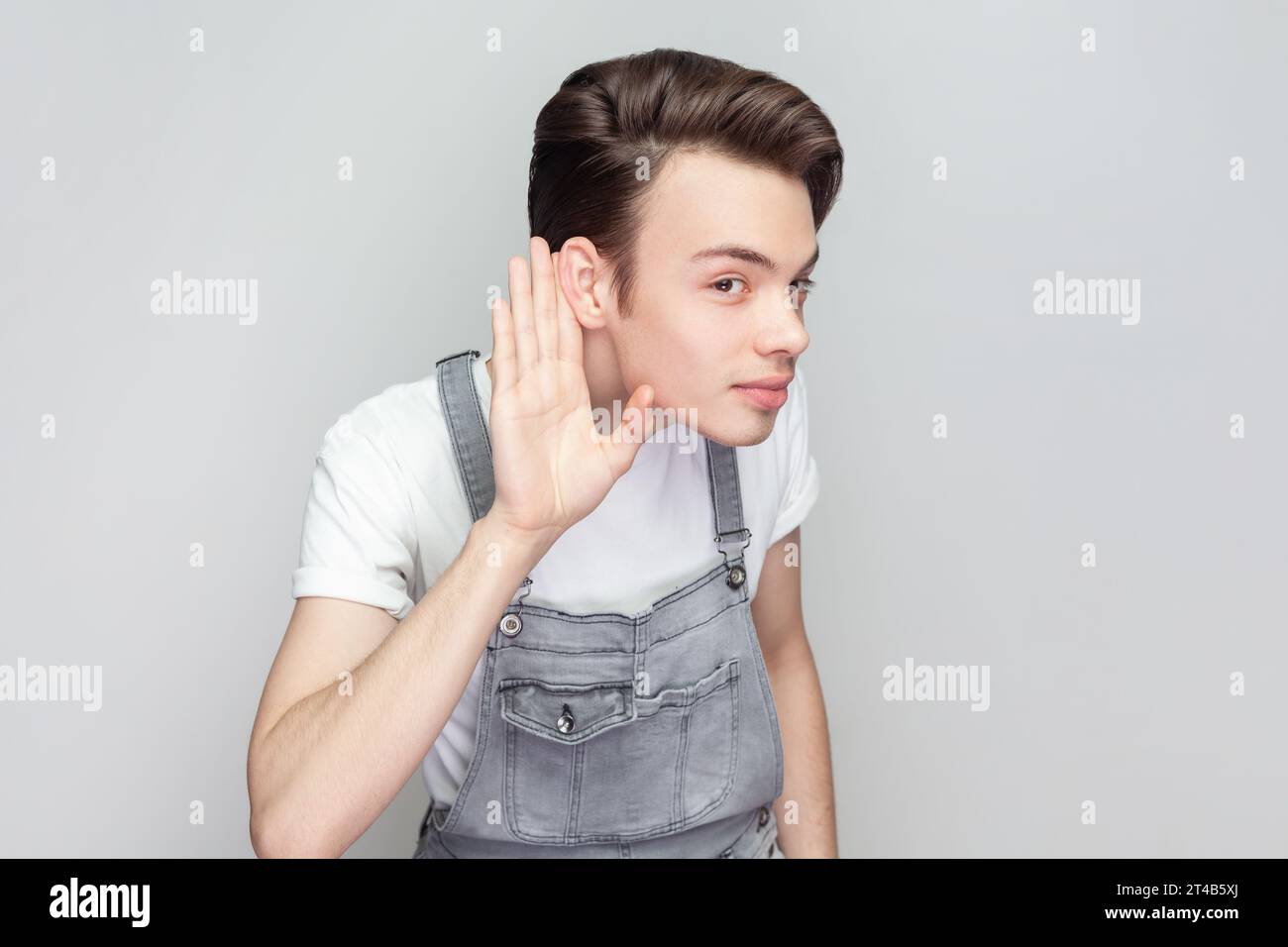 Intrigued curious young brunette man standing keeps hand near ear tries ...