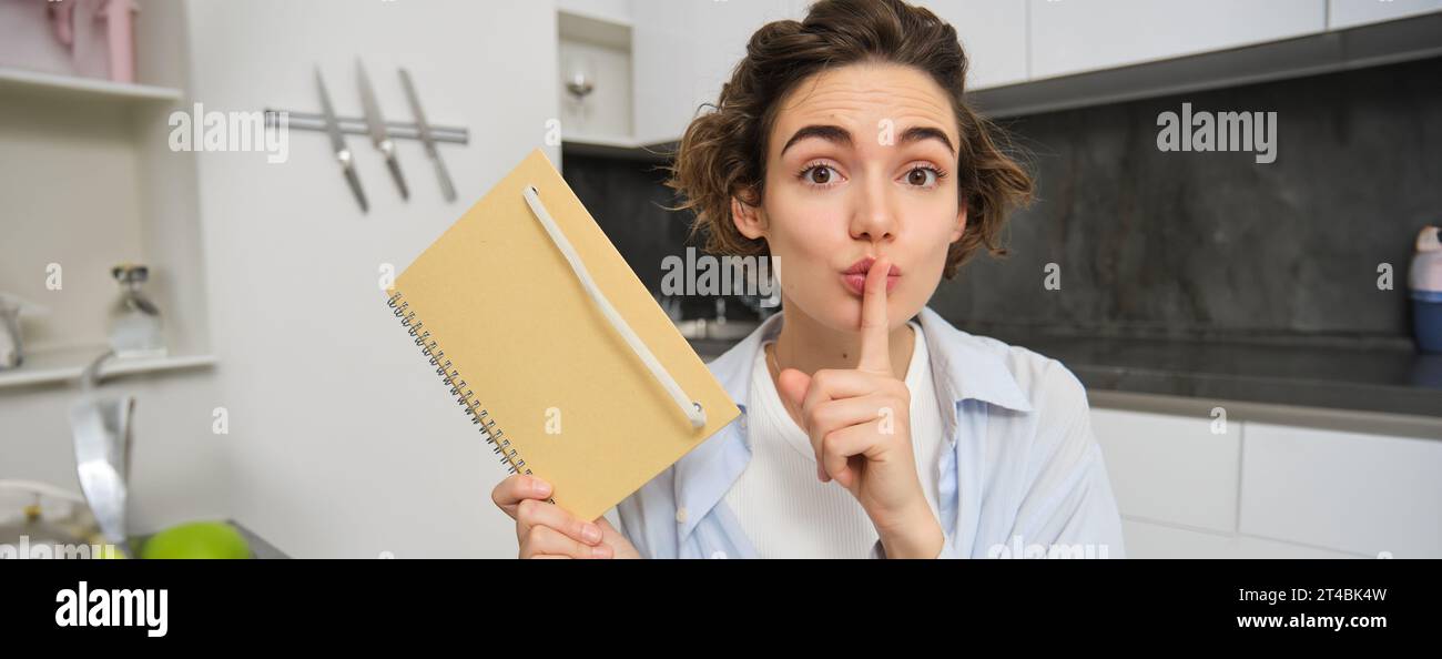 Young woman holds her diary, shows secret, hush gesture, presses finger ...