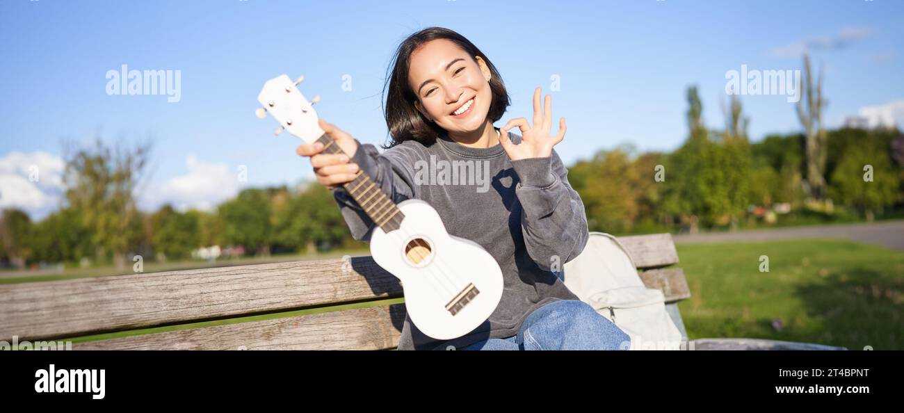 Cute smiling girl shows ok sign and her new ukulele, sits on bench in ...