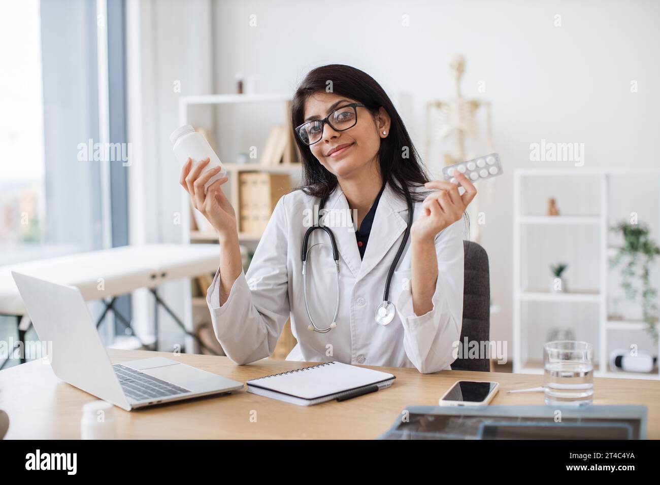 Optimistic lady doctor sitting at workplace and holding medicine for ...