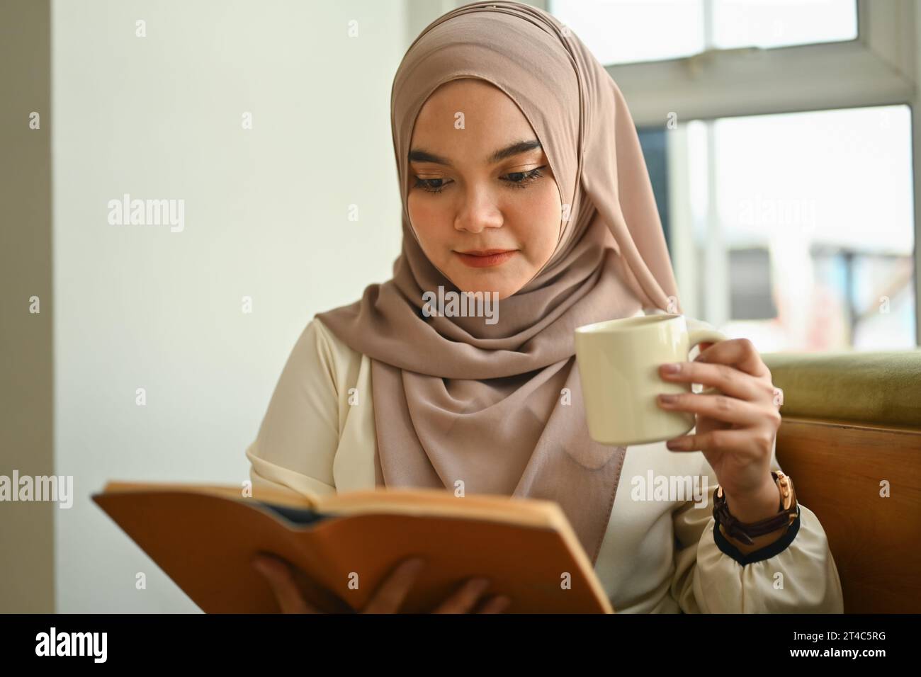 Relaxed muslim woman drinking hot tea and reading book in coffee shop ...