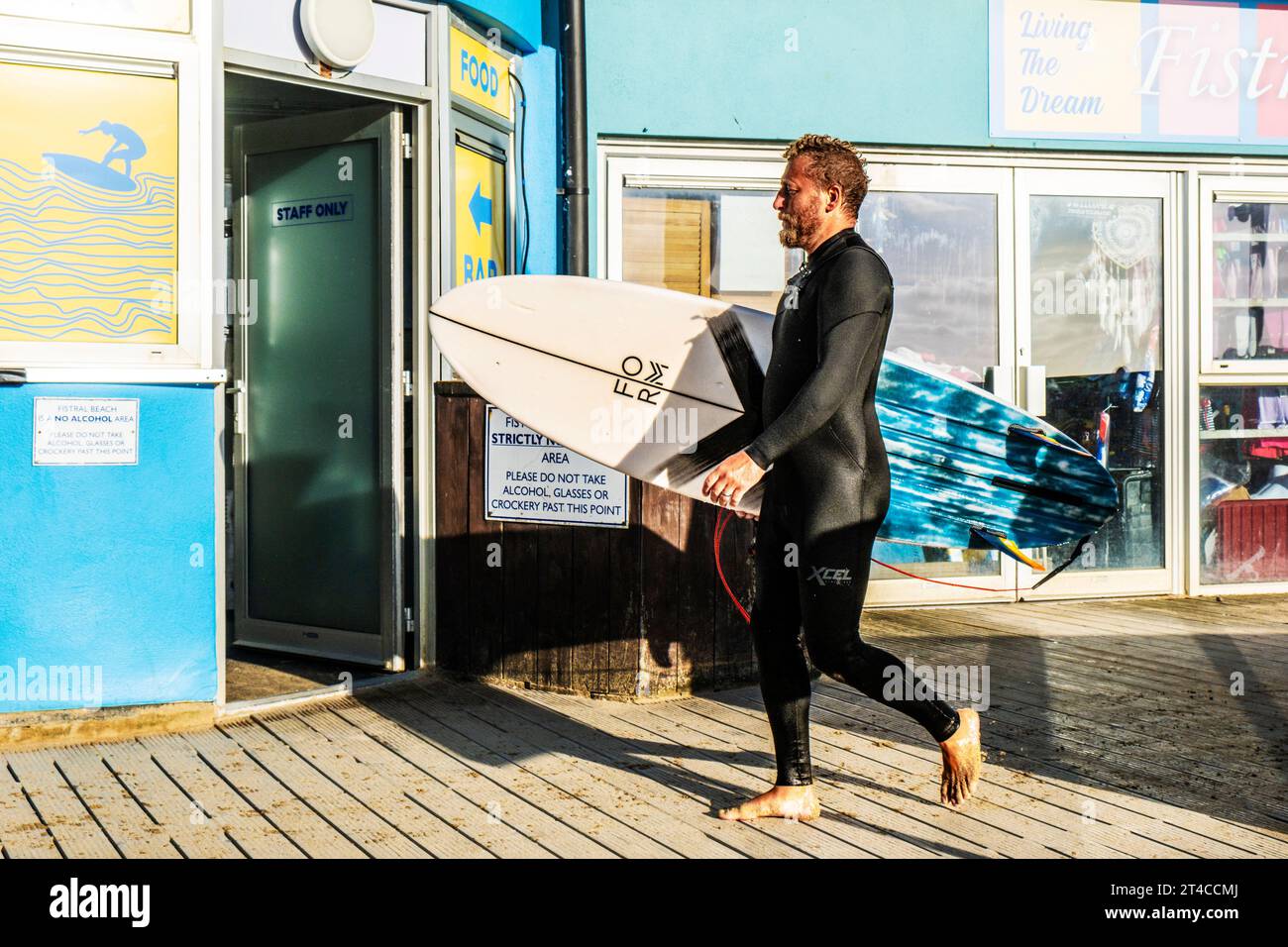 A male surfer carrying a Form surfboard walking past the entrance to ...