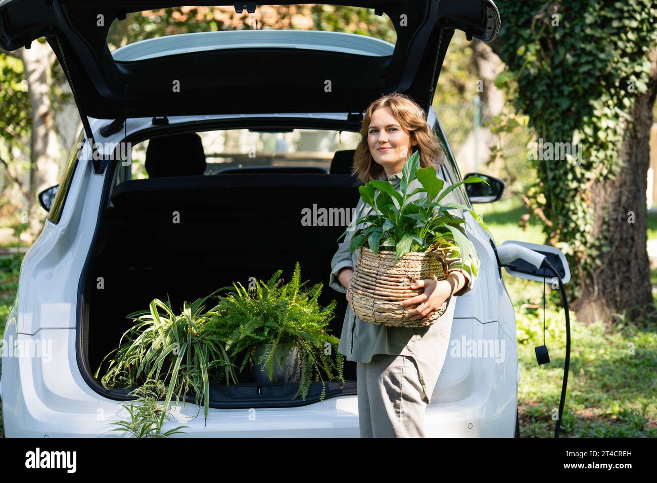 Woman with plant in pot next to a charging electric car in the yard of ...